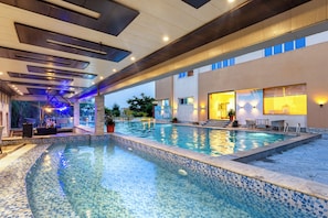 Piscina com borda azulejada, área coberta com assentos e um edifício com uma grande janela.