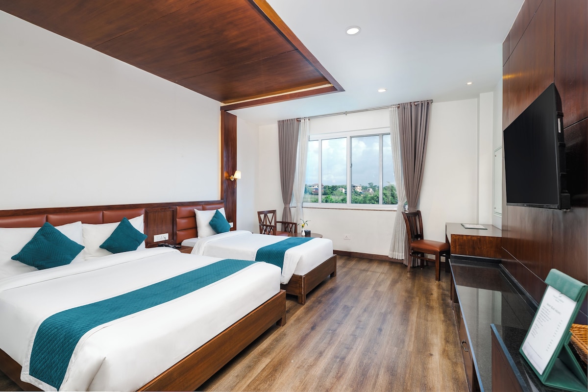 Quarto de hotel com duas camas, uma escrivaninha, televisão e janela com cortinas.