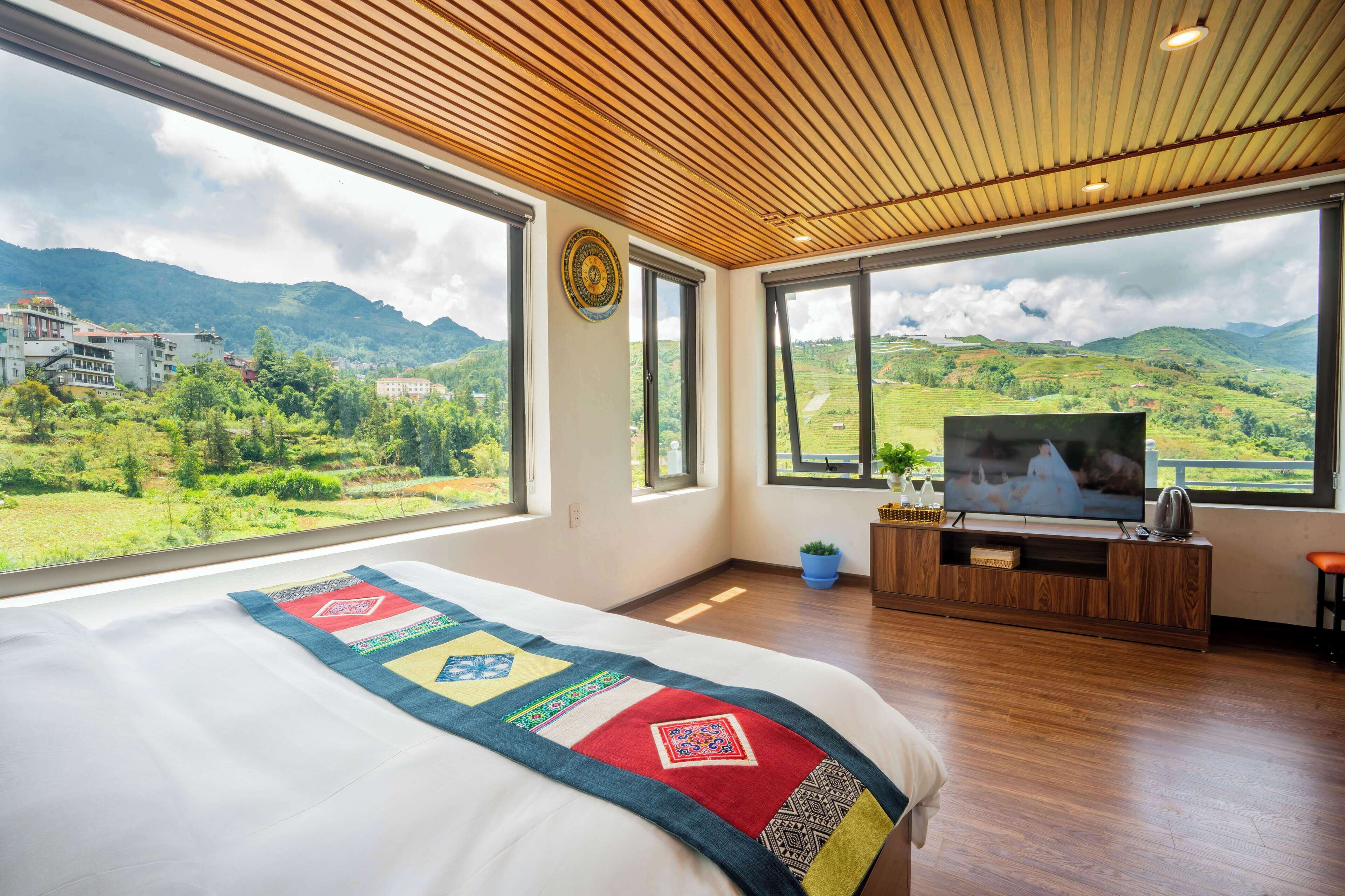 Quarto com uma cama grande, televisão e vista para uma paisagem montanhosa através de amplas janelas.