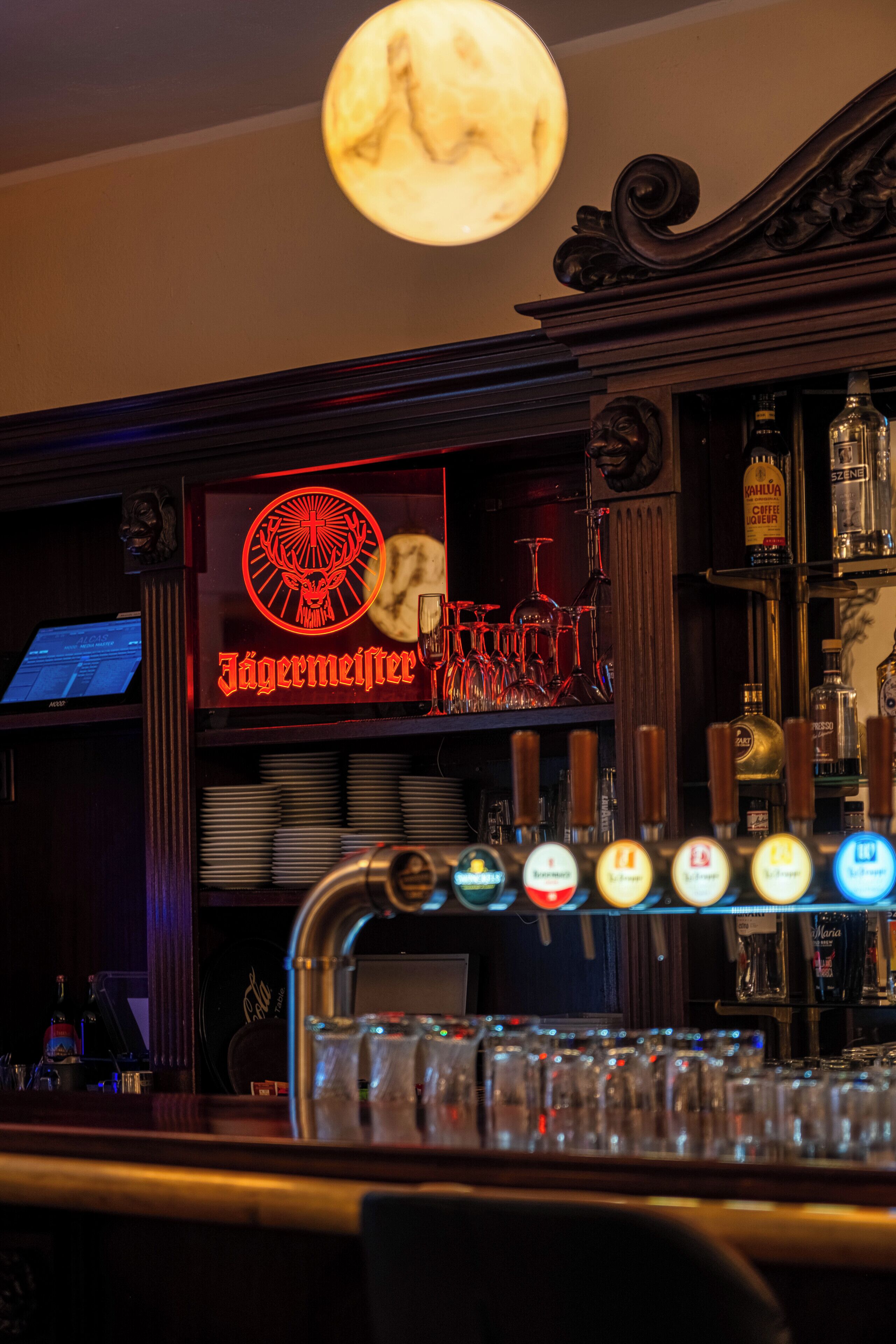 Uma área de bar com um letreiro de neon que diz "Jägermeister", uma seleção de garrafas, copos e um balcão.