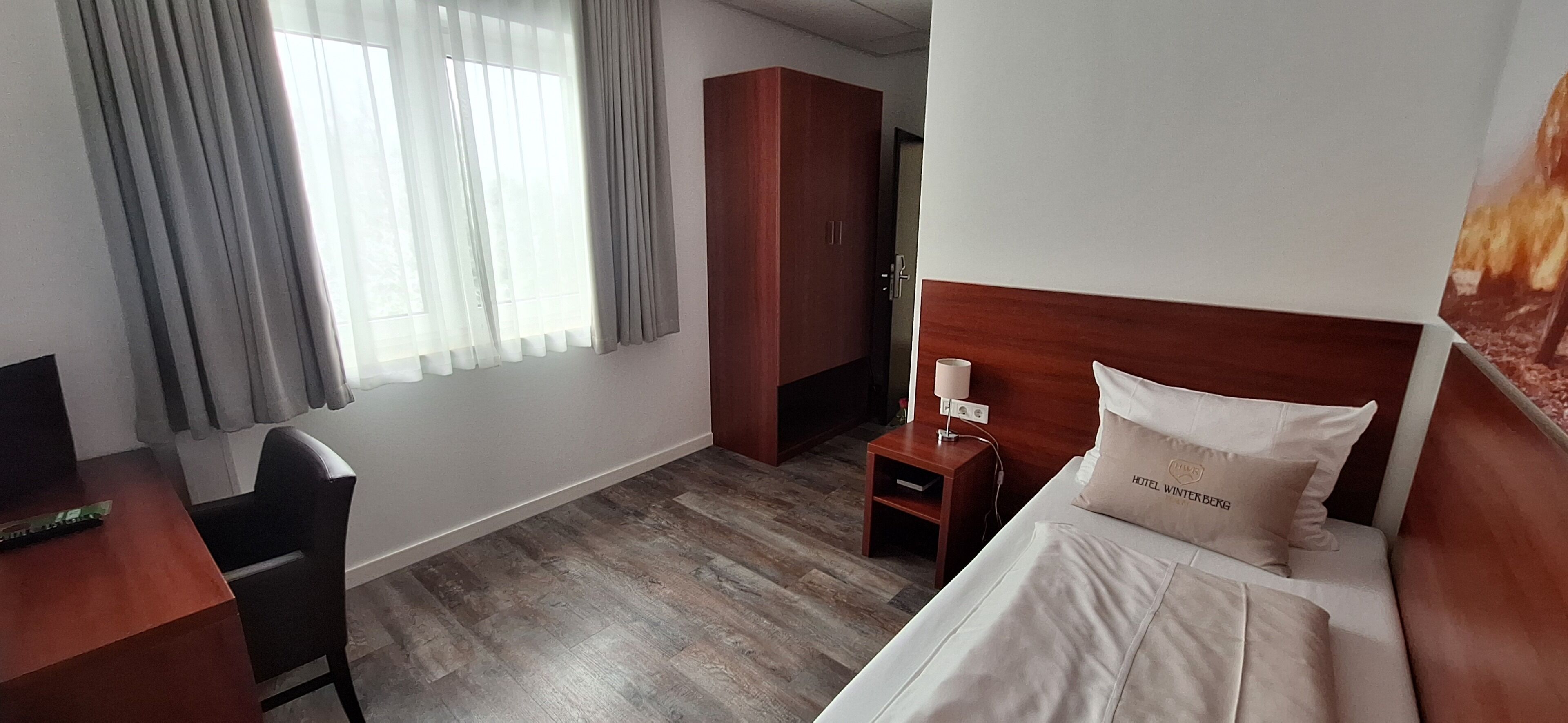 Quarto de hotel com cama, escrivaninha, cadeira, guarda-roupa e janela com cortinas.