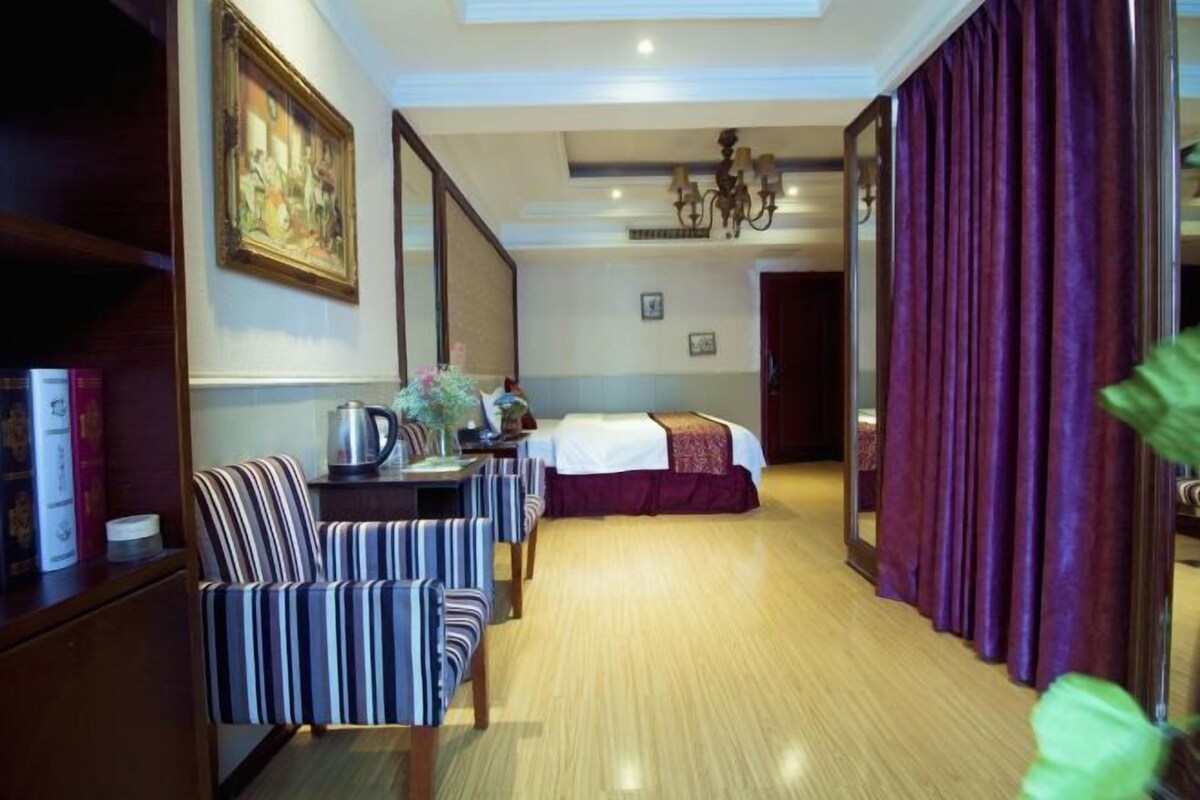 Quarto de hotel com piso de madeira, cama com colcha branca e vermelha, poltrona listrada, uma pequena mesa com chaleira e um quadro na parede.