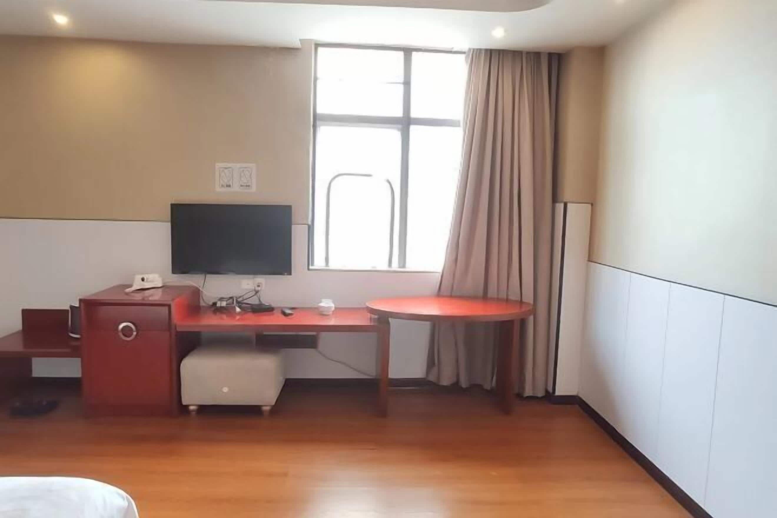 Quarto de hotel com uma escrivaninha de madeira, televisão, janela com cortinas e uma cama.