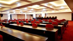 Uma sala de conferência com fileiras de mesas e cadeiras, um palco e piso coberto por um tapete vermelho.