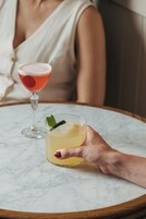 Uma mão segurando um drink com folha de hortelã sobre uma mesa de mármore.
