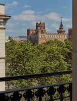 Vista de um castelo histórico com várias torres e uma torre do relógio imponente, cercado por árvores verdes exuberantes.