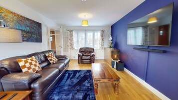 Uma sala de estar com sofá de couro, mesa de centro de madeira, tapete azul, poltrona de couro marrom e televisão fixada na parede.