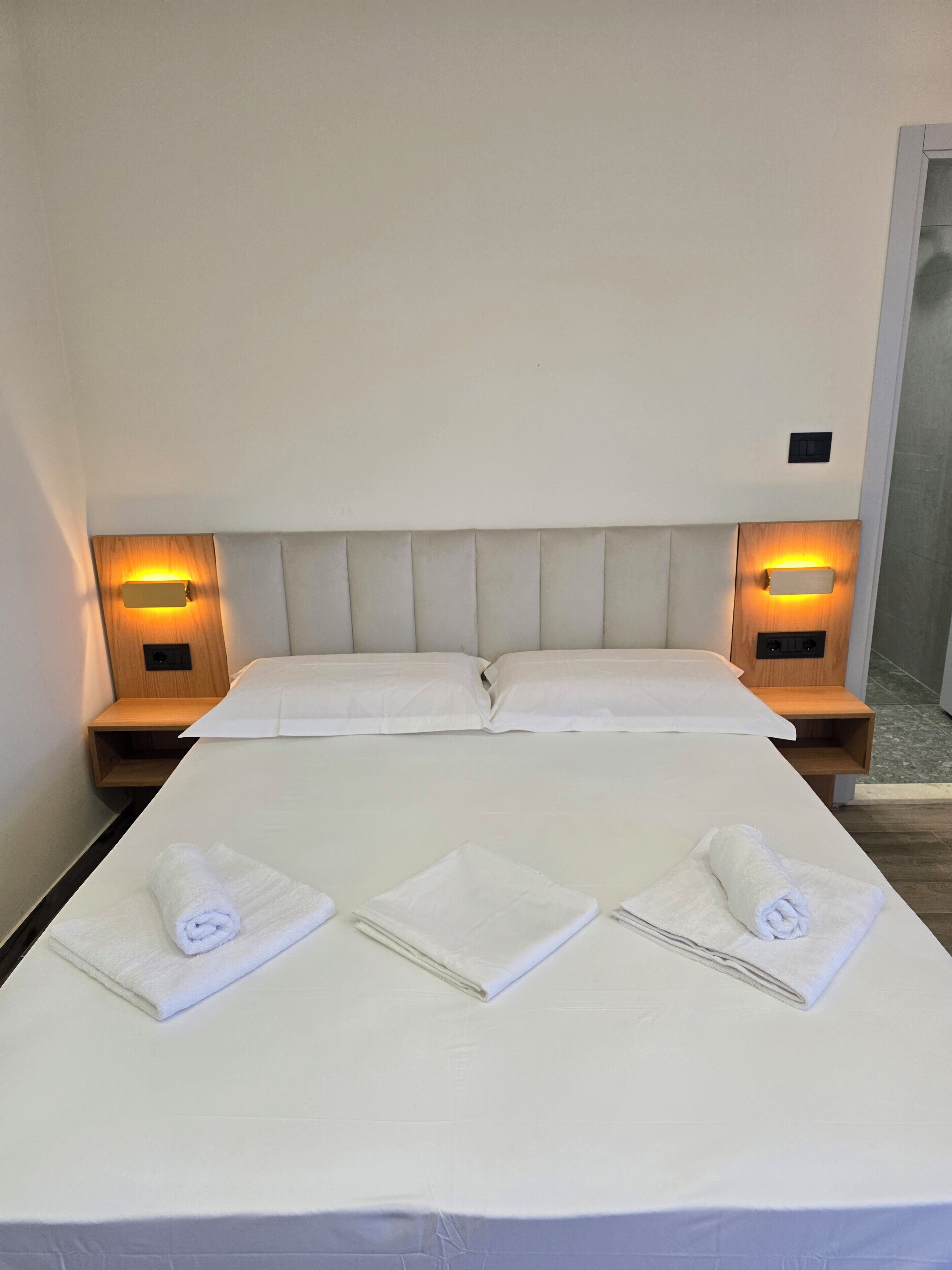 Quarto de hotel com uma cama grande, duas mesas de cabeceira e luminárias fixadas na parede.