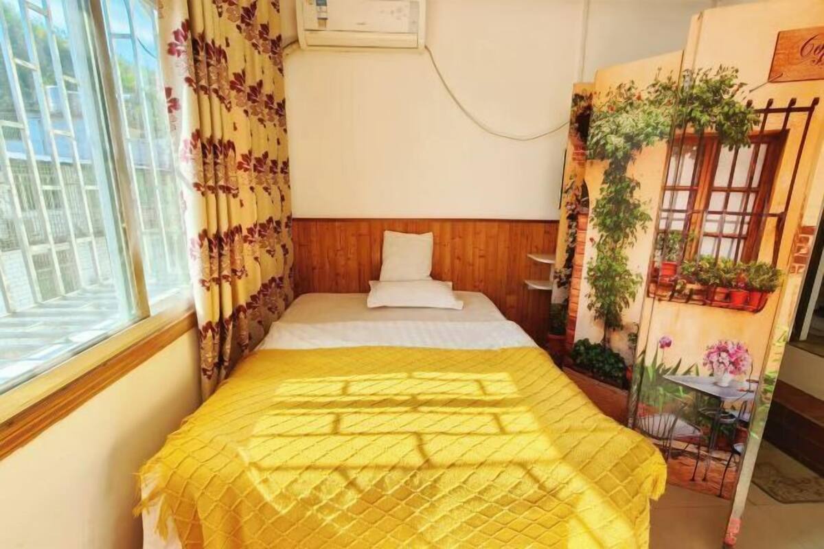 Uma cama com colcha amarela, uma janela com cortinas e uma parede com arranjo de plantas.