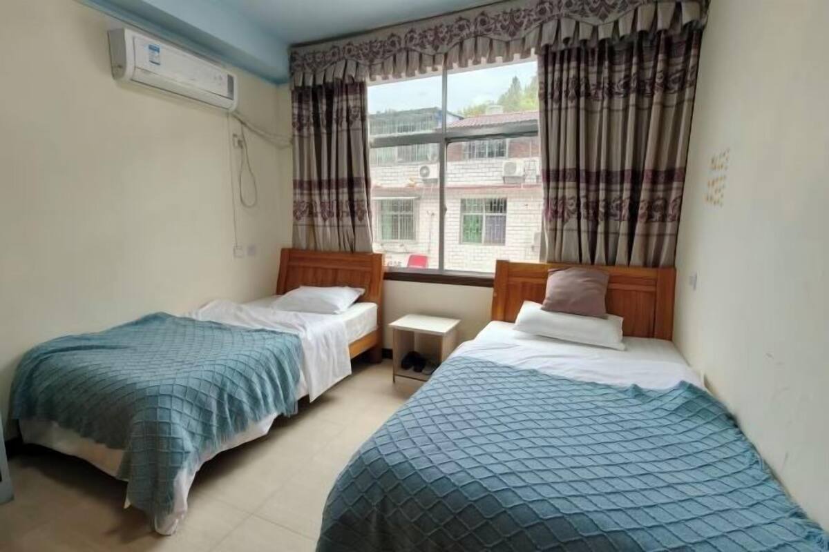 Quarto com duas camas, uma janela com cortinas, uma mesinha de cabeceira pequena e um ar condicionado.