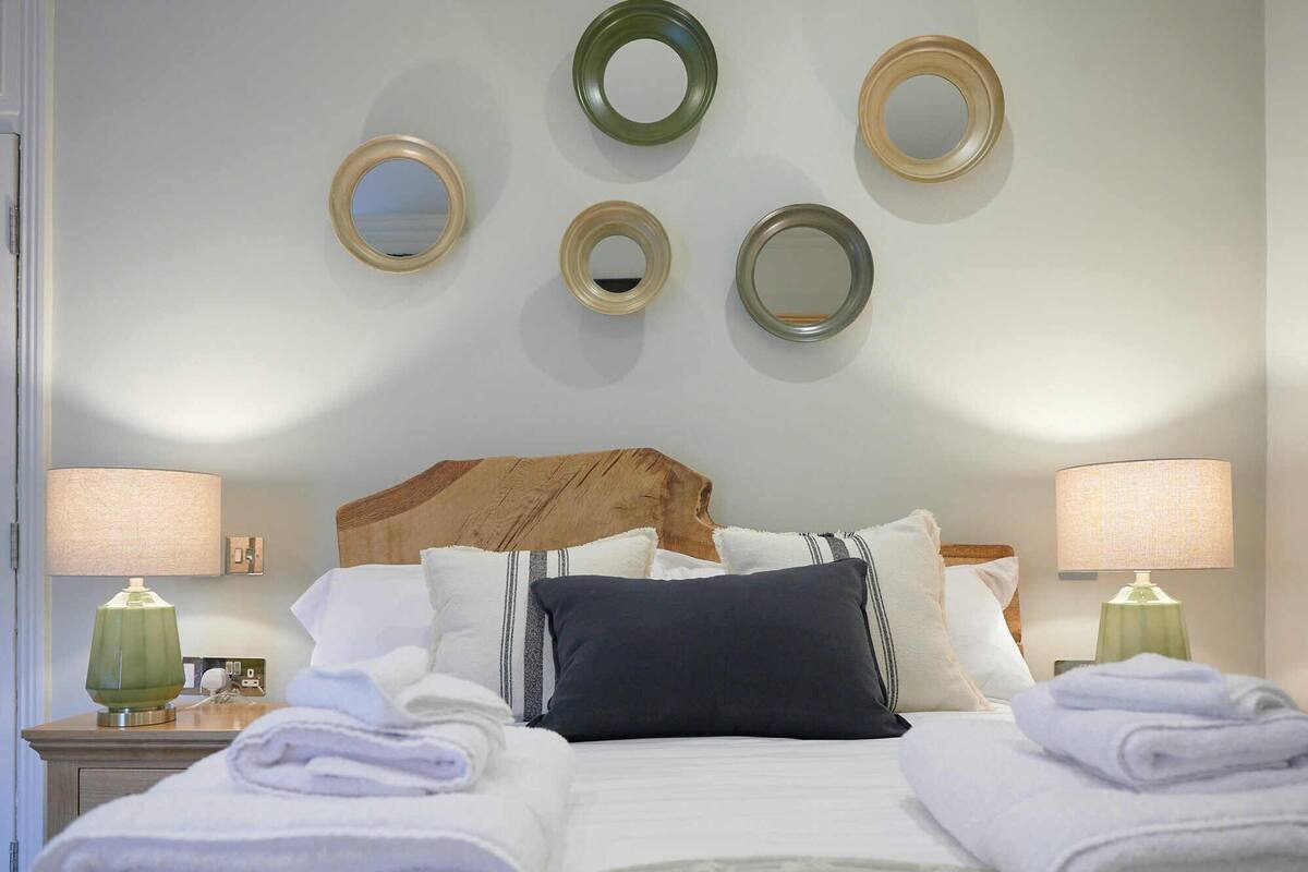 Quarto de hotel com cabeceira de madeira, cama com roupa de cama branca e listrada, duas luminárias de apoio e quatro espelhos fixados na parede.