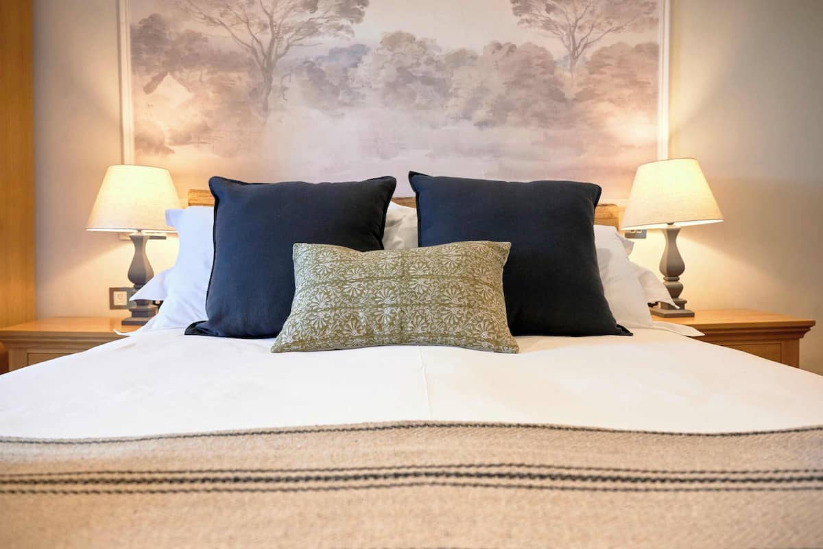 Cama bem arrumada com duas almofadas escuras e uma almofada estampada, com duas luminárias de cabeceira em mesas de cabeceira de madeira.