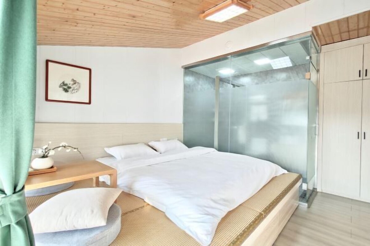 Quarto moderno com cama, mesa de cabeceira, quadro emoldurado e área de chuveiro com divisória de vidro.