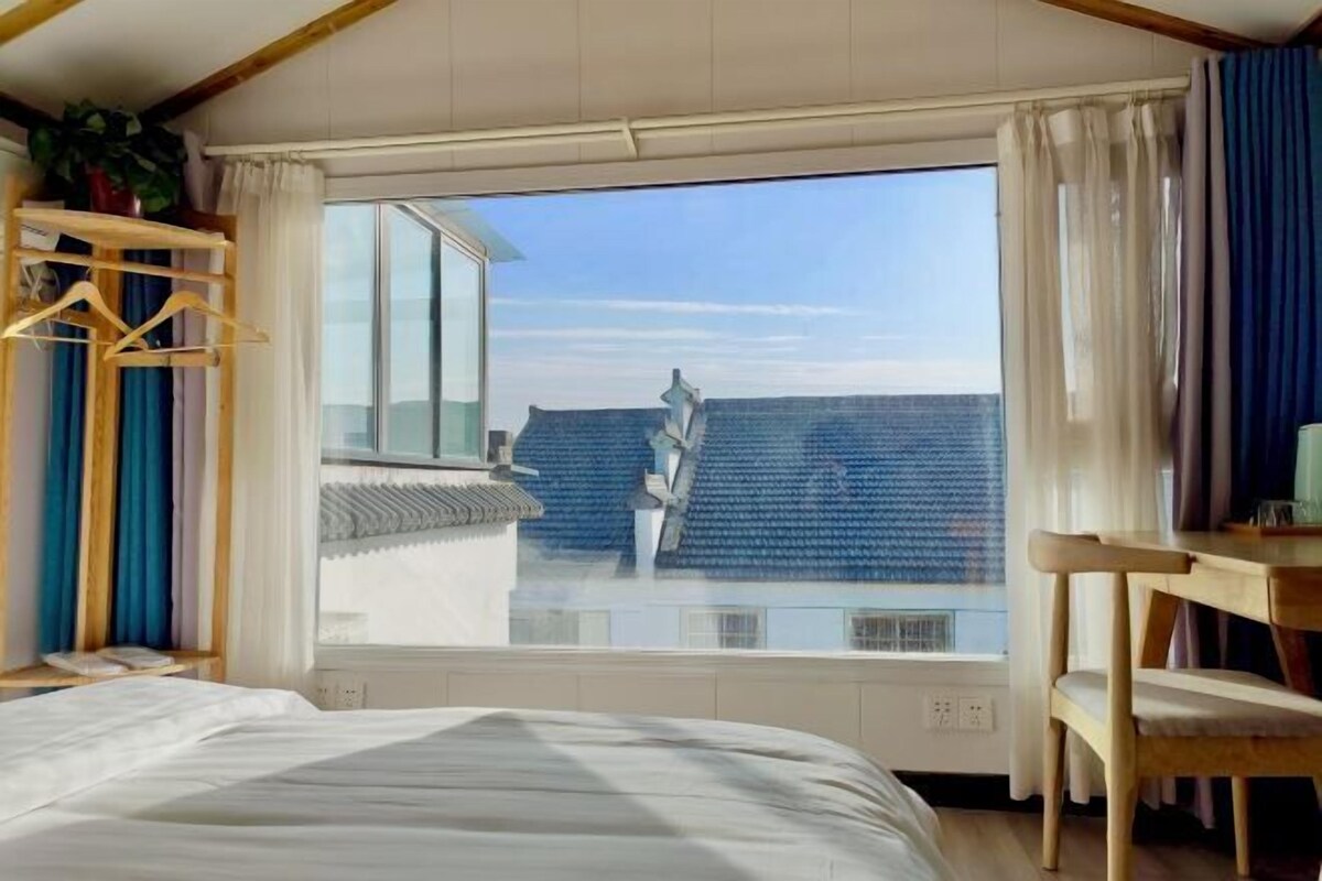 Quarto com uma janela grande, uma cama, uma cadeira de madeira e uma planta.