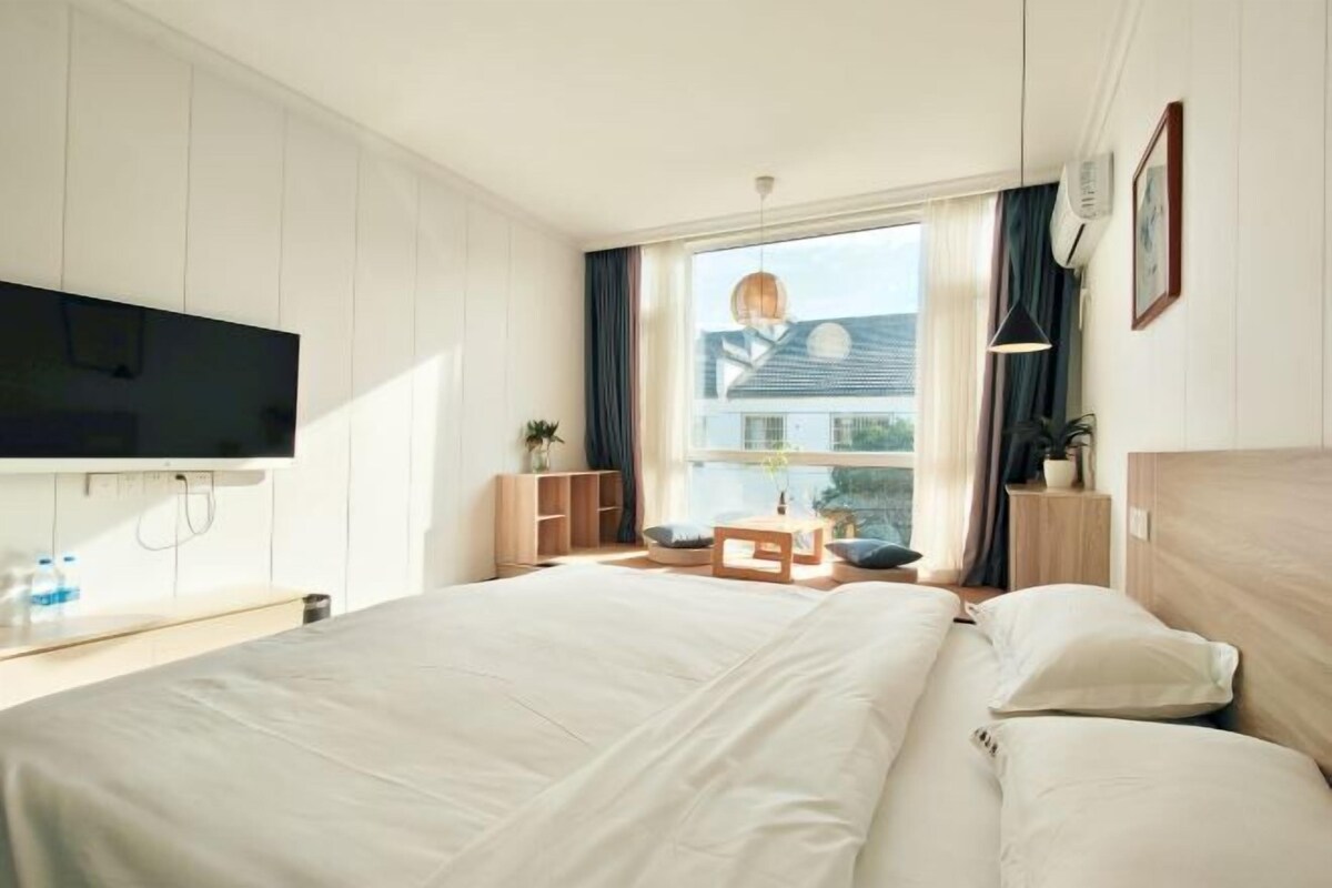 Quarto de hotel com uma cama grande, televisão na parede, um pequeno espaço de estar e uma janela com cortinas.