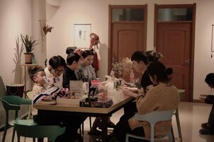 Um grupo de pessoas sentado à volta de uma mesa num quarto, com uma porta e uma planta ao fundo.