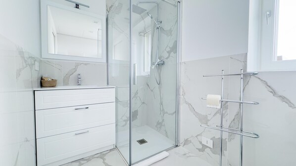 Um banheiro moderno com chuveiro em box de vidro, toucador branco com gavetas e parede revestida de mármore.