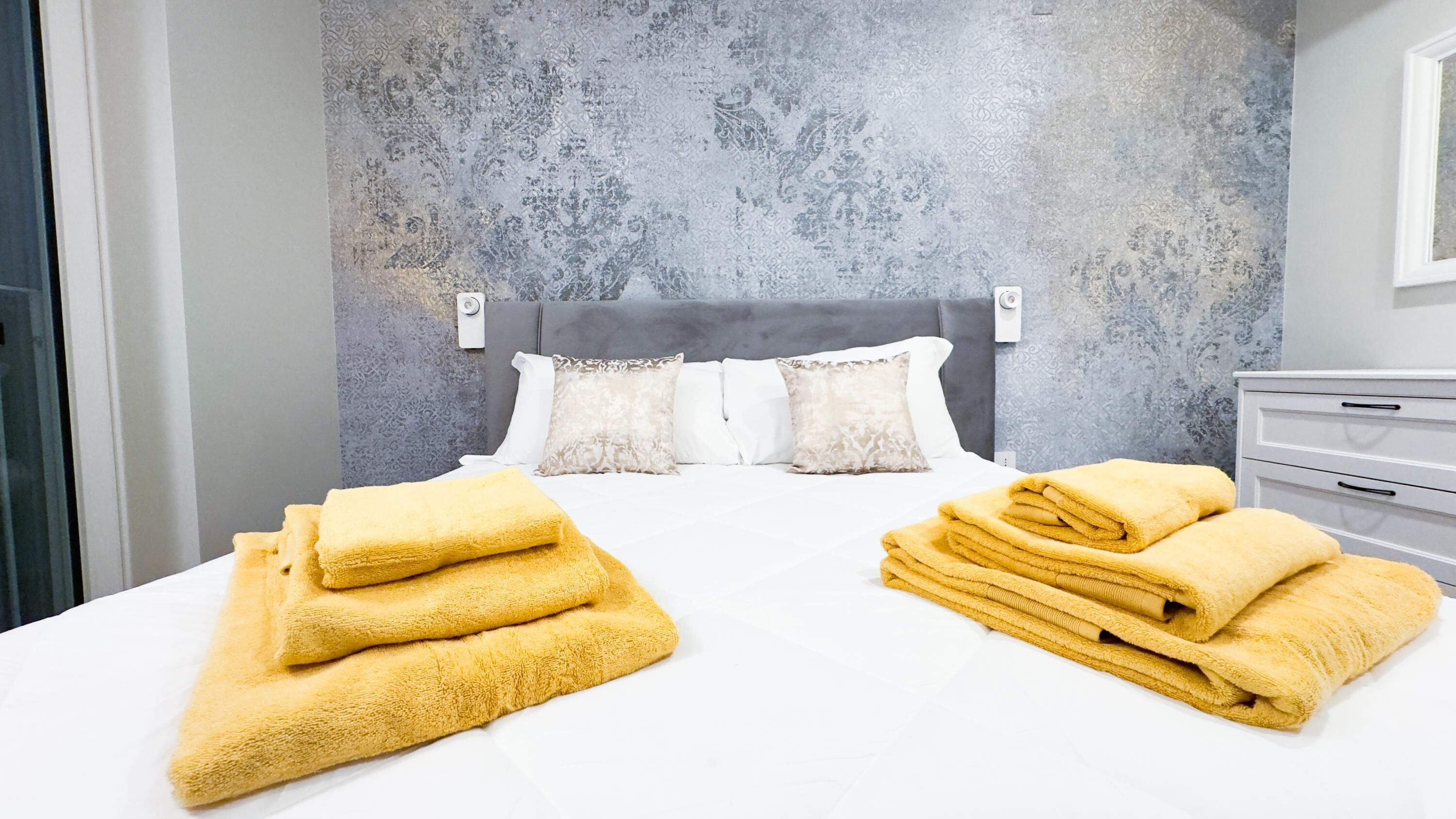 Cama bem arrumada com duas pilhas de toalhas amarelas, parede texturizada cinza e mesa de cabeceira branca com gavetas.