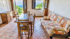 Uma sala de jantar com mesa e cadeiras de madeira, sofá estampado e vista para o exterior através de janelas abertas.