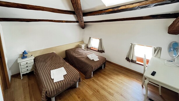 Quarto com duas camas, vigas de madeira, uma pequena escrivaninha e uma janela com cortinas.