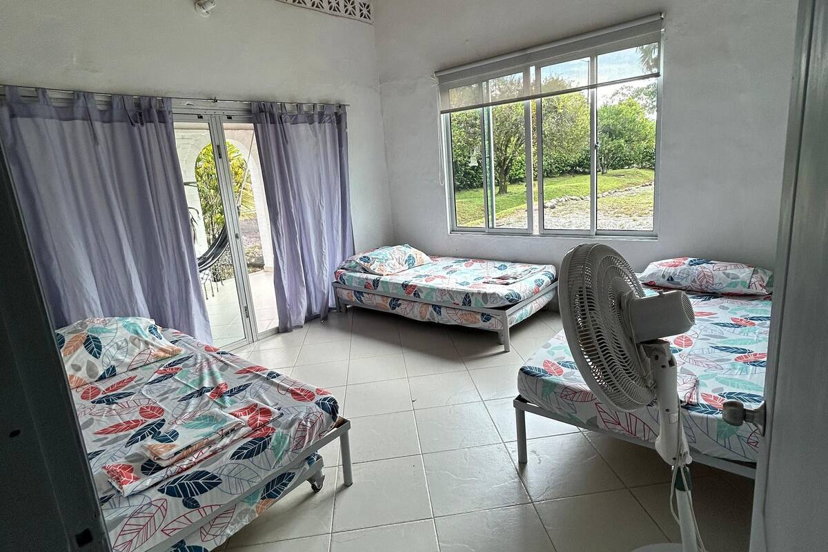 Quarto com três camas, ventilador de teto e janela com vista para o verde.