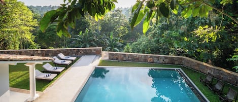 Piscina retangular com borda de pedra, rodeada por um muro de pedra e uma pérgula branca.