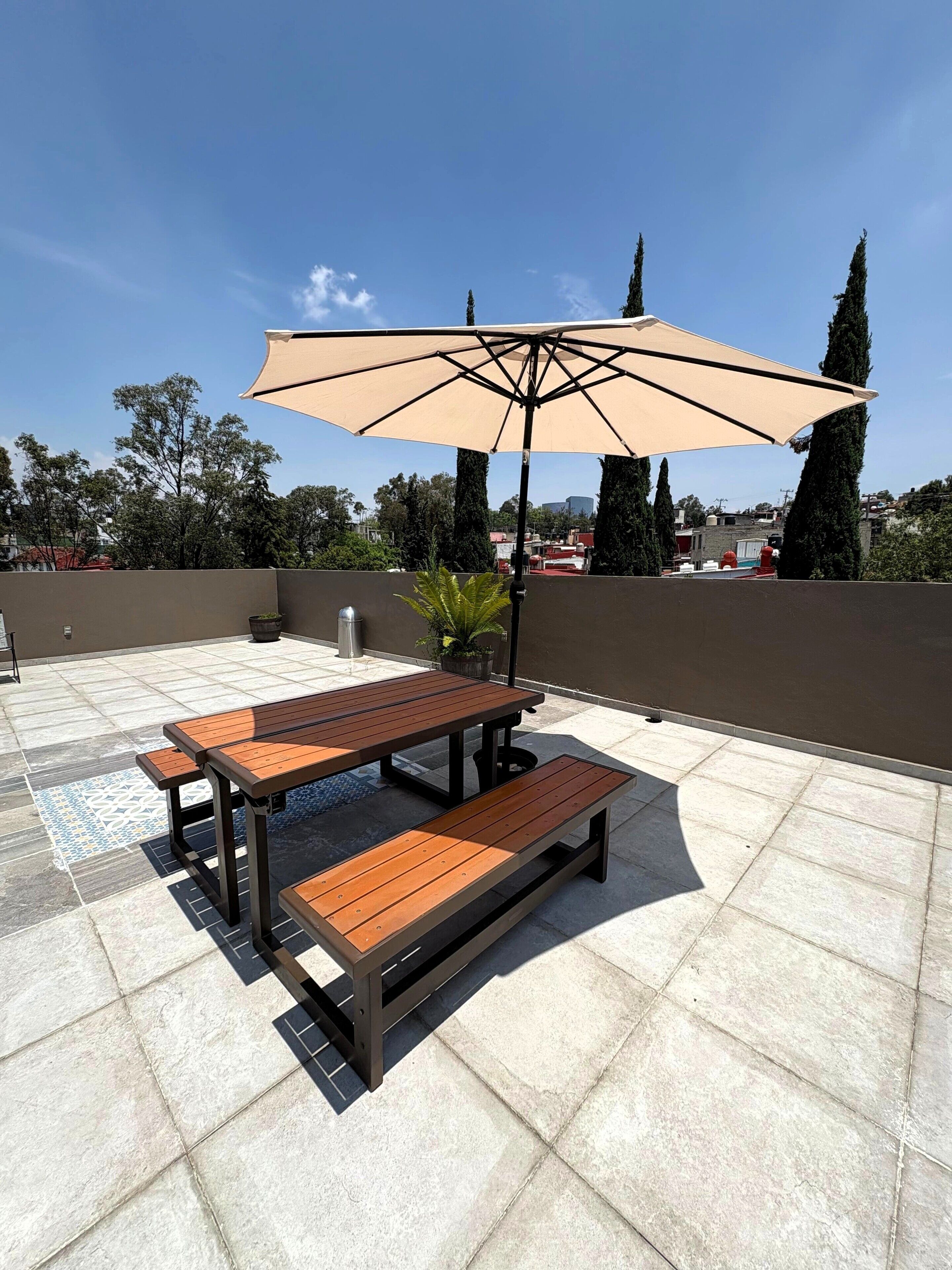Um terraço no telhado com mesa e bancos de madeira, um grande guarda-sol bege e vista para a paisagem urbana.