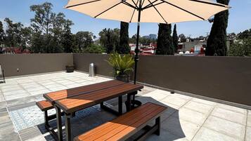 Um terraço no telhado com mesa e bancos de madeira, um grande guarda-sol bege e vista para a paisagem urbana.