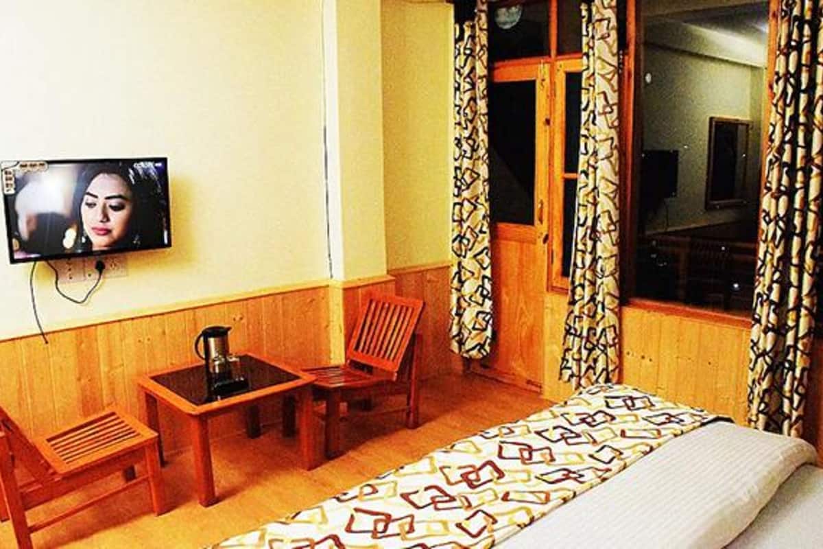 Quarto de hotel com cama, televisão na parede, mesa pequena com cadeira e cortinas na janela.