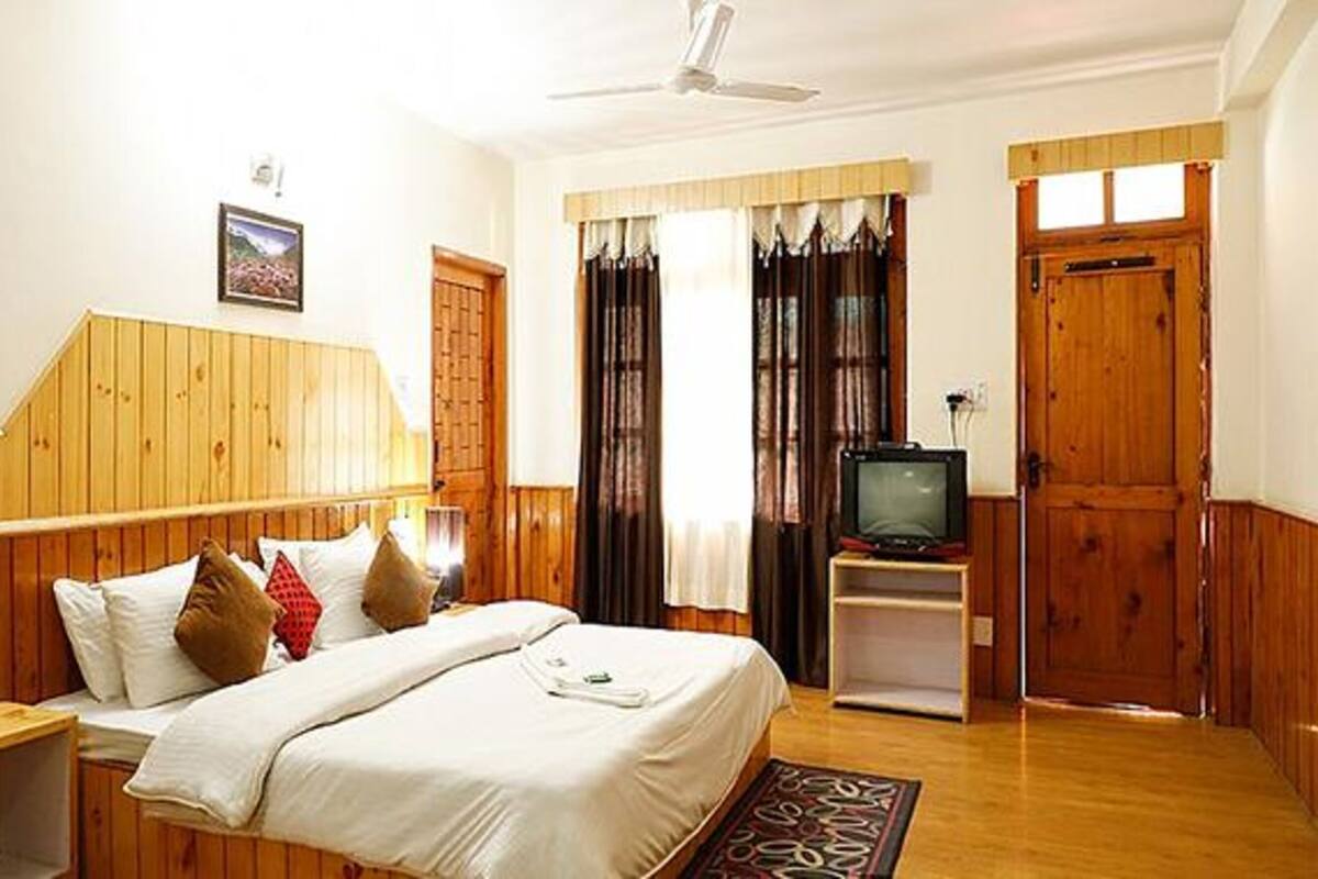 Quarto com cabeceira de madeira, cama com roupa de cama branca, televisão sobre um suporte e janela com cortinas.