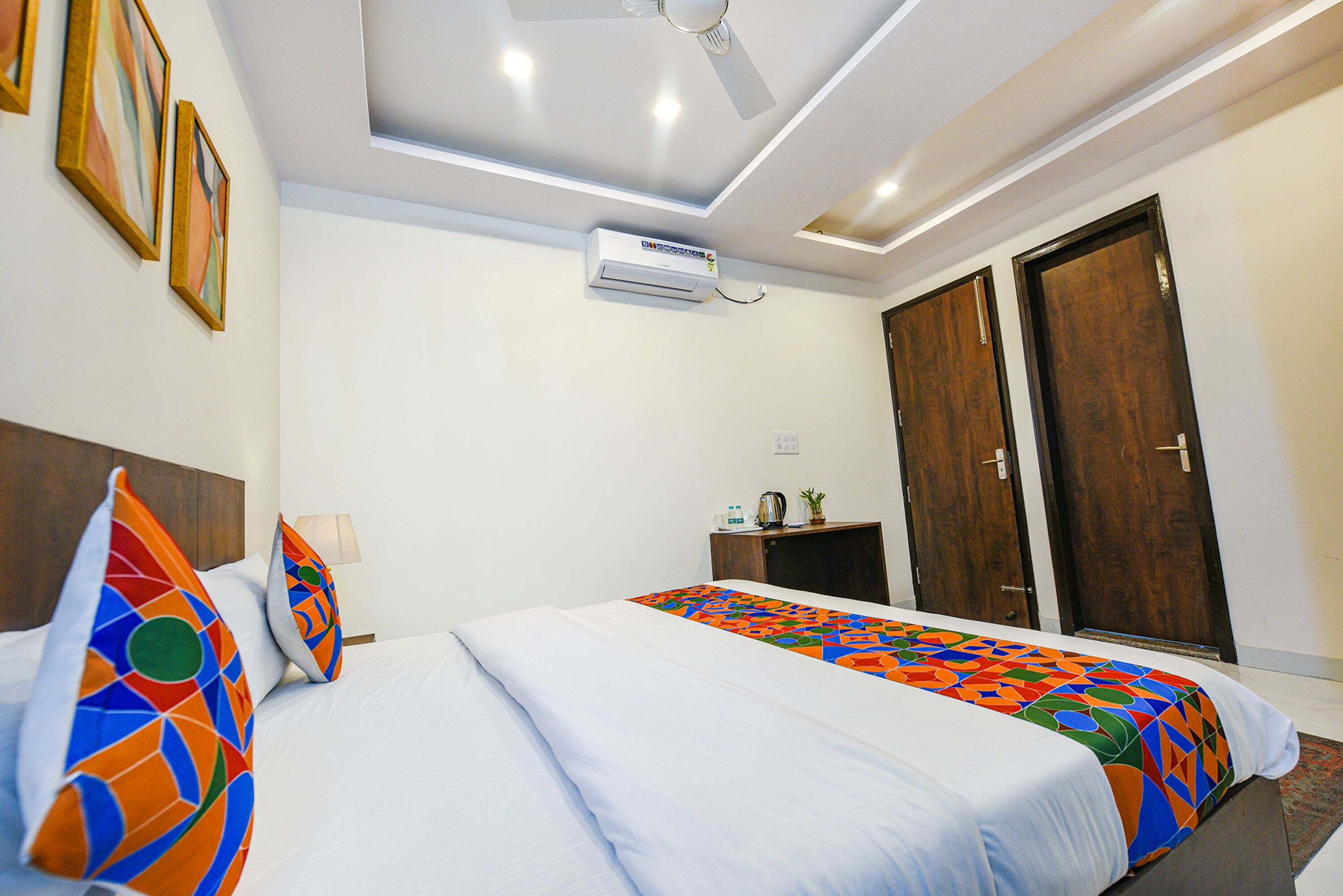 Quarto de hotel com cama, ventoinha no teto, aparelho de ar condicionado, mesa de cabeceira, luminária e guarda-roupa.