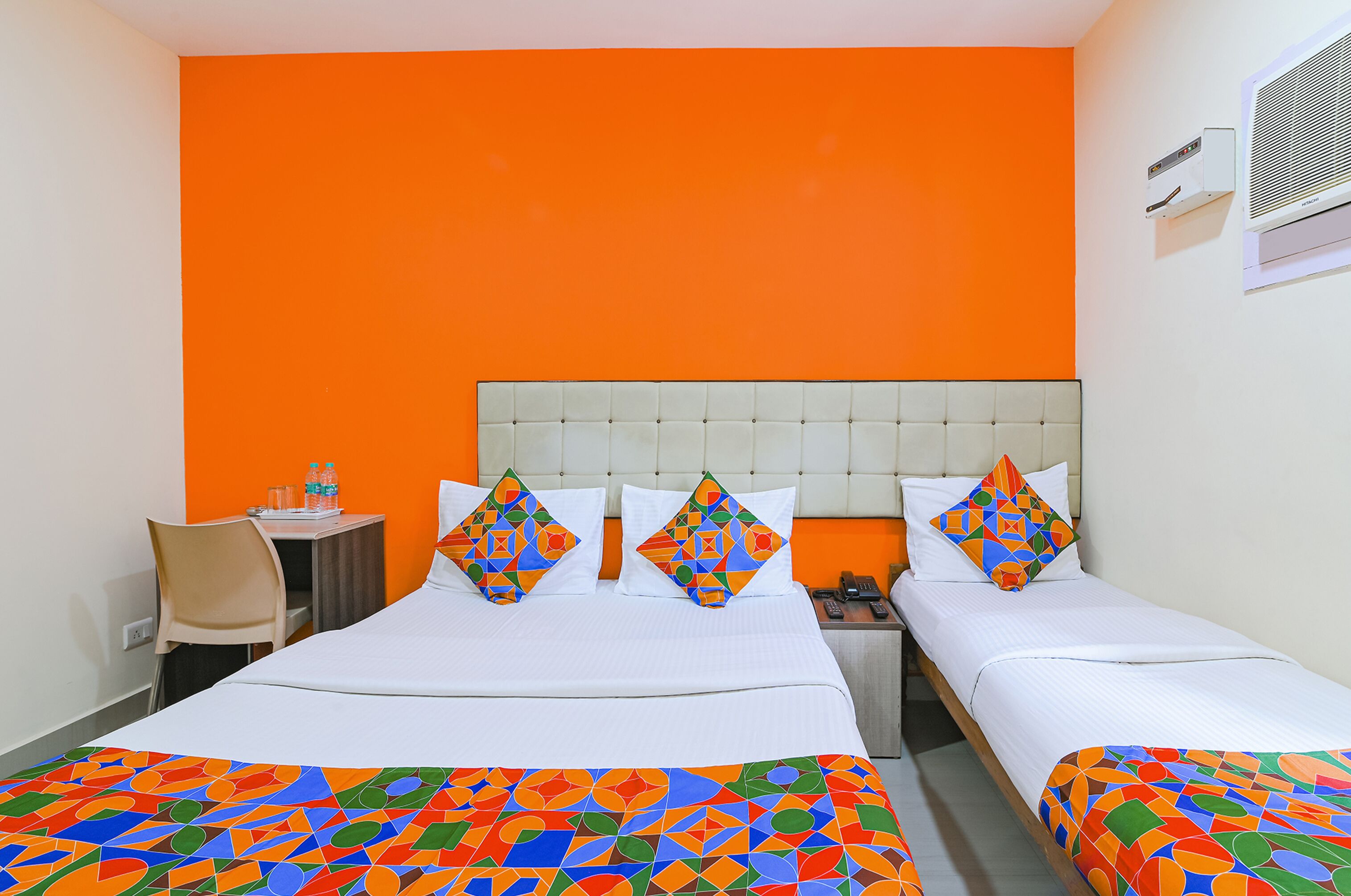 Quarto de hotel com duas camas, uma mesa pequena, uma cadeira e um aparelho de ar condicionado na parede.