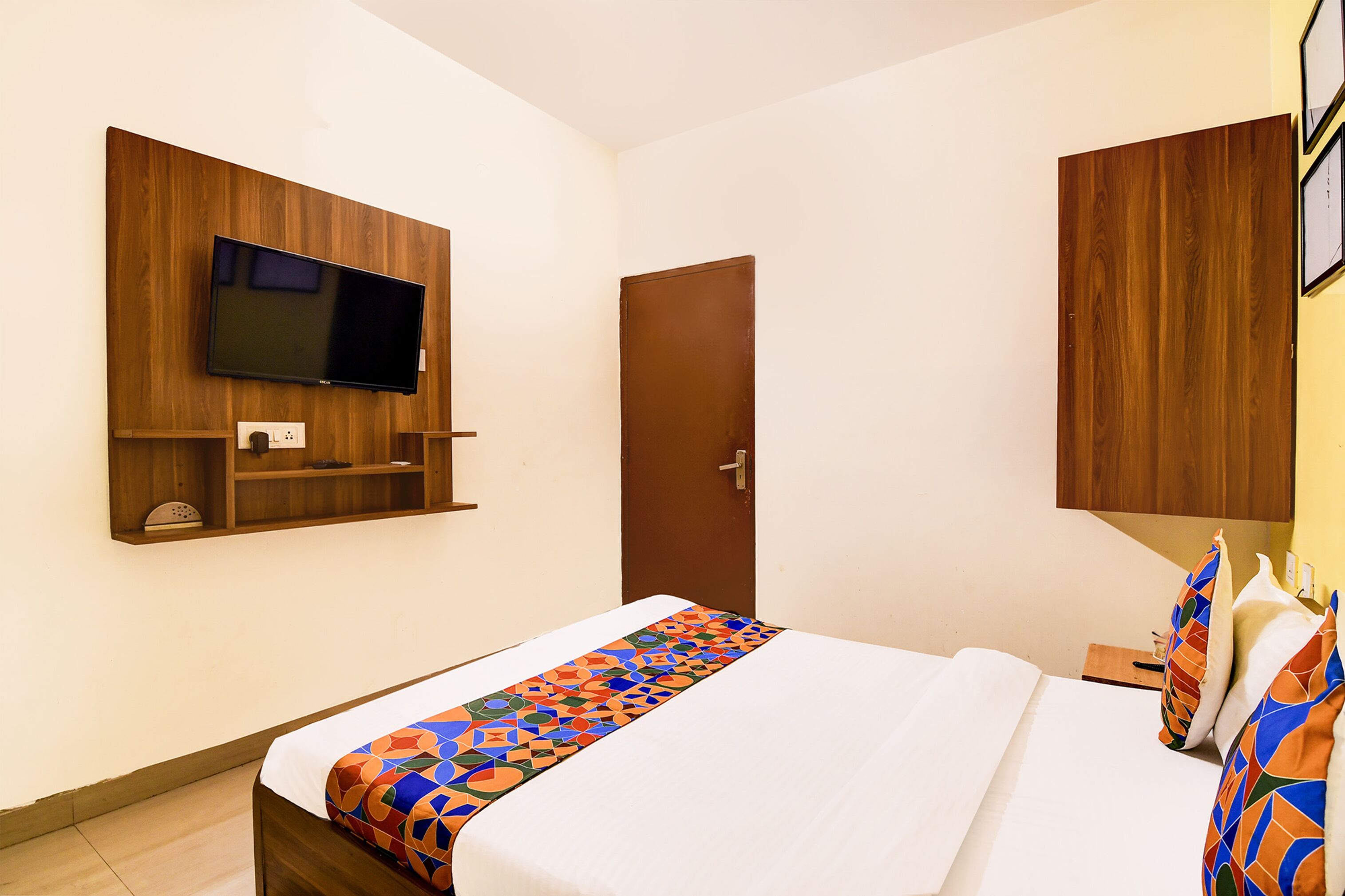 Quarto de hotel com cama, televisão fixada na parede, guarda-roupa de madeira e uma porta.