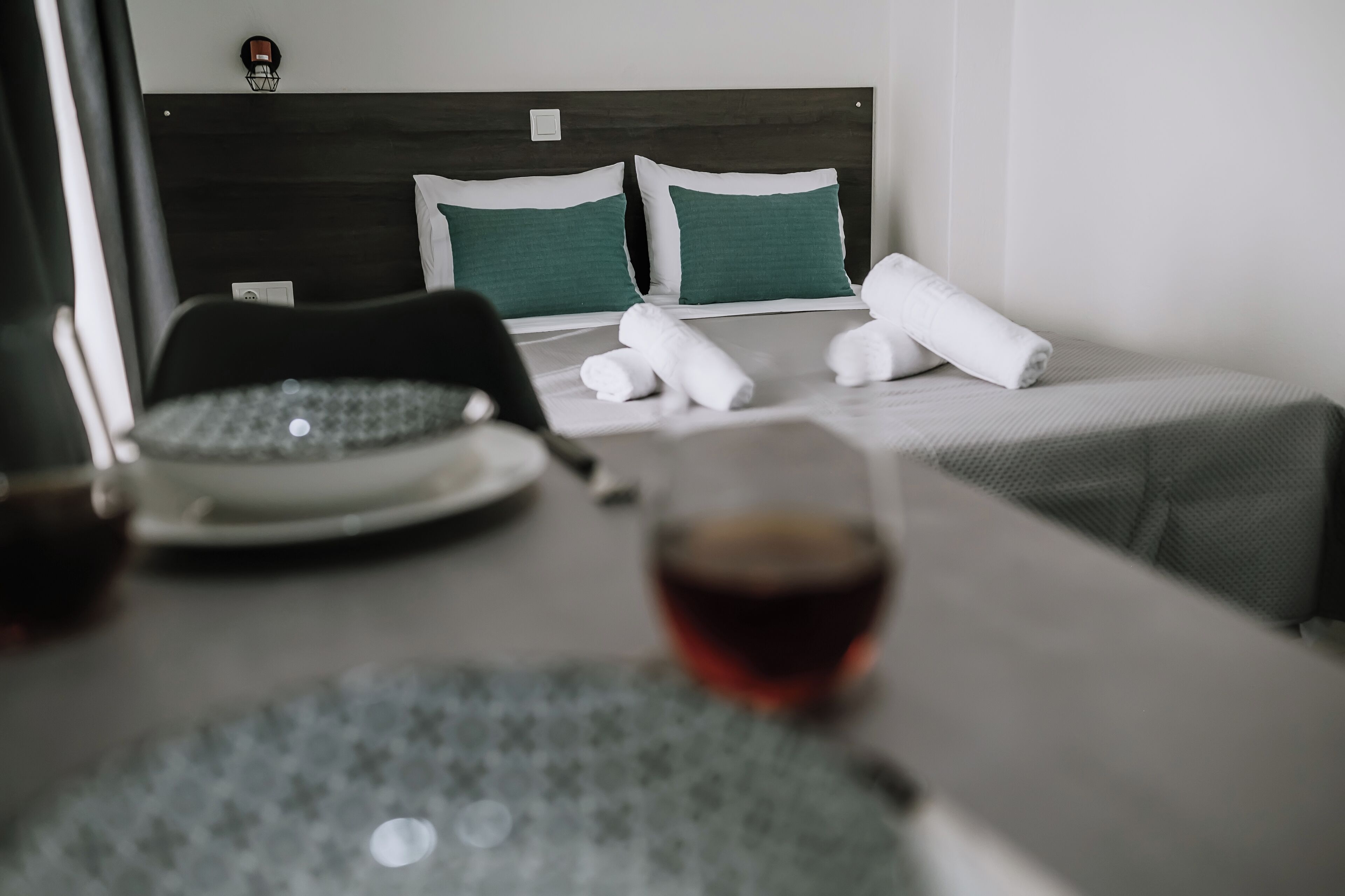 Quarto de hotel bem arrumado com cama, almofadas, uma mesa com um copo de vinho tinto e uma cadeira.
