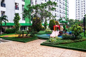 Um parquinho com escorrega e estruturas de escalada, cercado por vegetação bem cuidada e um caminho de azulejos.