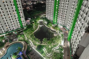 Um edifício alto com um pátio ajardinado, apresentando um lago e palmeiras, cercado por caminhos iluminados.