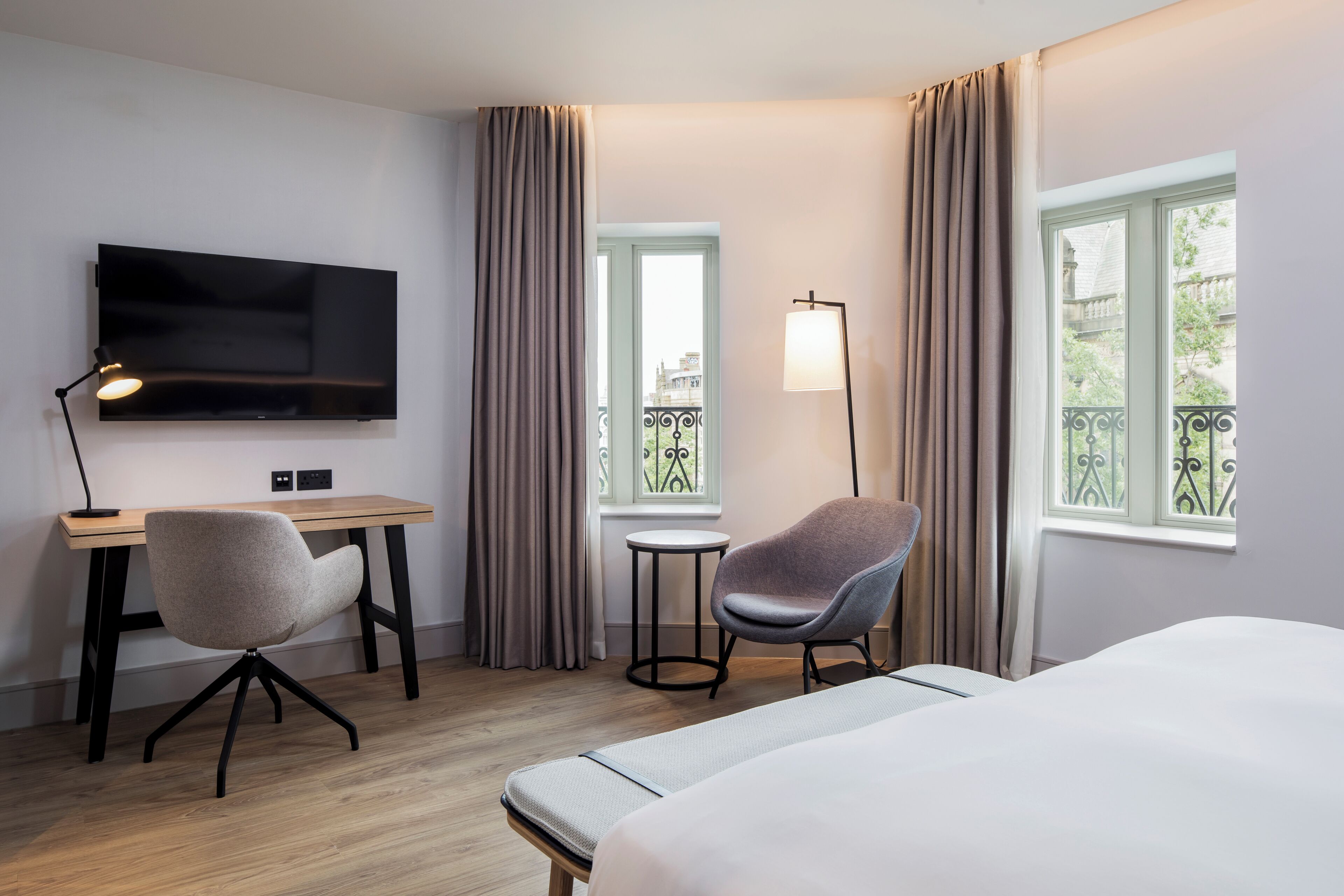 Quarto de hotel com cama, escrivaninha, cadeira, televisão e janela com cortinas.