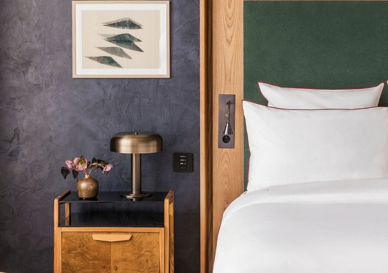 Quarto de hotel bem arrumado, com mesa de cabeceira de madeira, um criado-mudo com abajur, uma obra de arte emoldurada na parede e uma cama com roupa de cama branca.