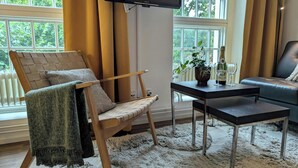 Uma sala de estar moderna com uma cadeira de madeira, uma mesa pequena, uma televisão e vista para o verde através de grandes janelas.