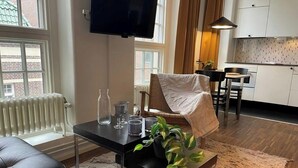 Uma sala de estar moderna com um sofá de couro preto, uma mesa de centro preta com copos e uma planta, uma TV montada na parede e vista para a cozinha e exterior através de grandes janelas.