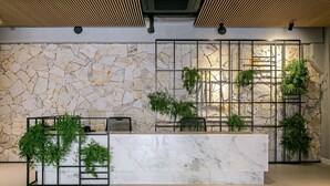 Uma receção com bancada de mármore, parede de pedra com padrão geométrico e plantas verdes em vasos de metal.