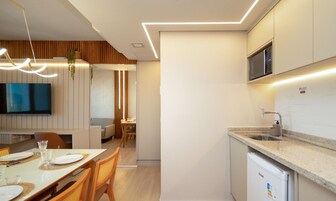 Cozinha moderna com micro-ondas embutido, pia e máquina de lavar louça. Há uma área de jantar com mesa e cadeiras, e uma televisão fixada na parede.
