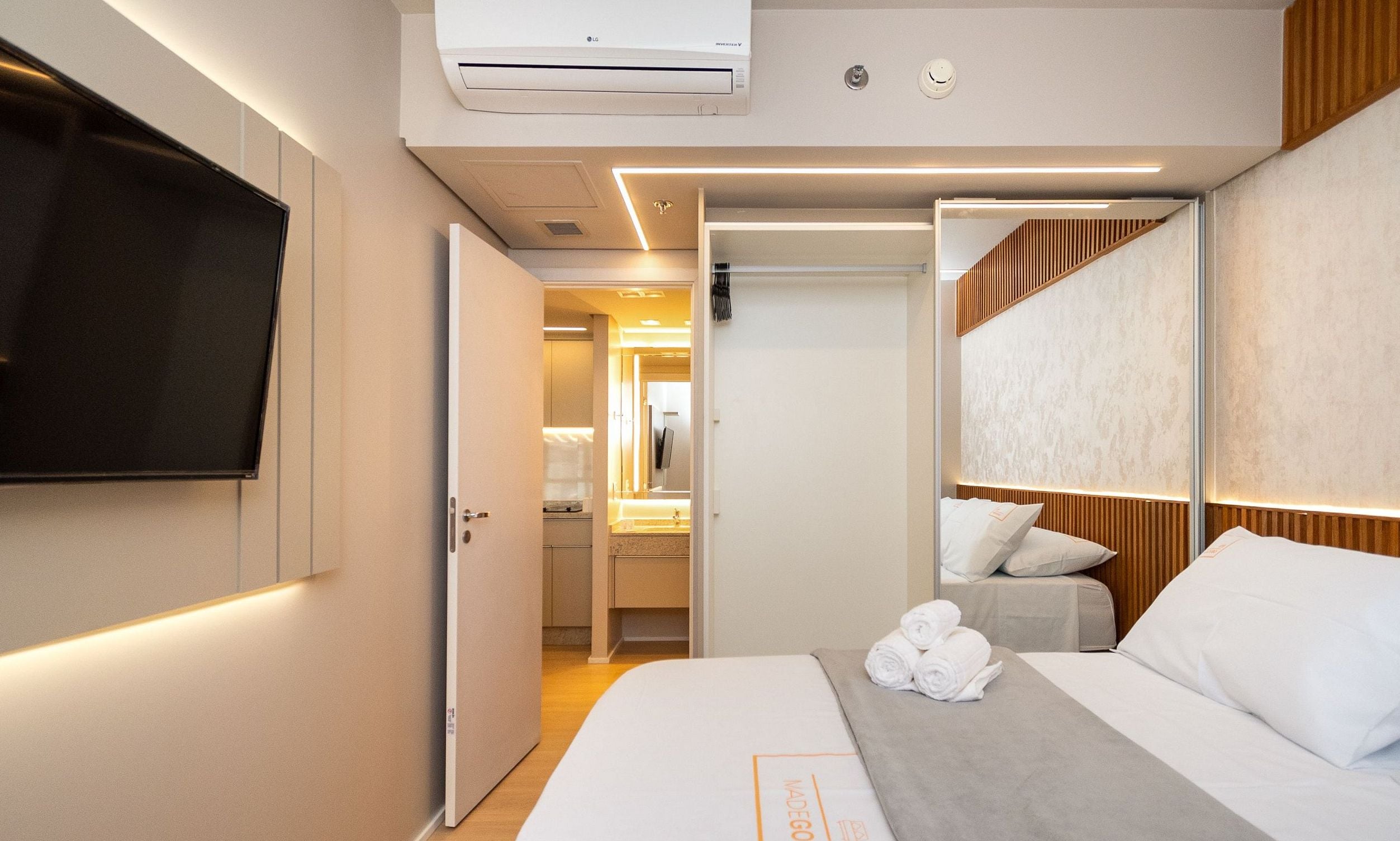 Quarto de hotel com cama, televisão, banheiro e corredor que dá para outro quarto.