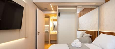 Quarto de hotel com cama, televisão, banheiro e corredor que dá para outro quarto.