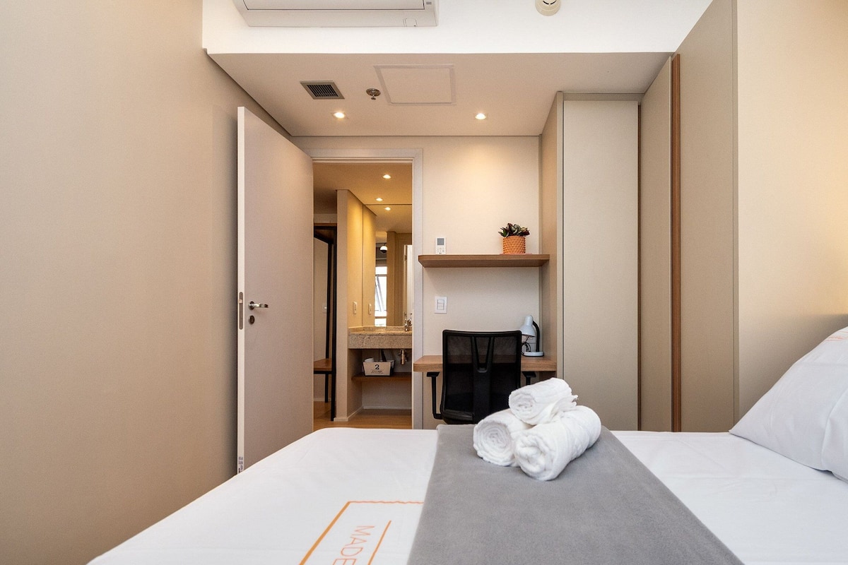 Quarto de hotel com uma cama, uma mesa com cadeira, uma pequena planta numa prateleira e um banheiro visível por uma porta aberta.
