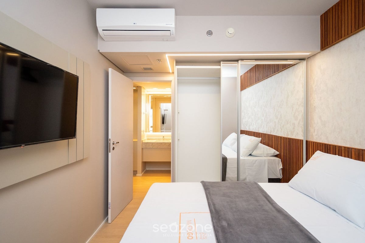 Quarto de hotel moderno com televisão de tela plana, cama com lençóis brancos, manta cinza, cabeceira de madeira e closet com espelho.