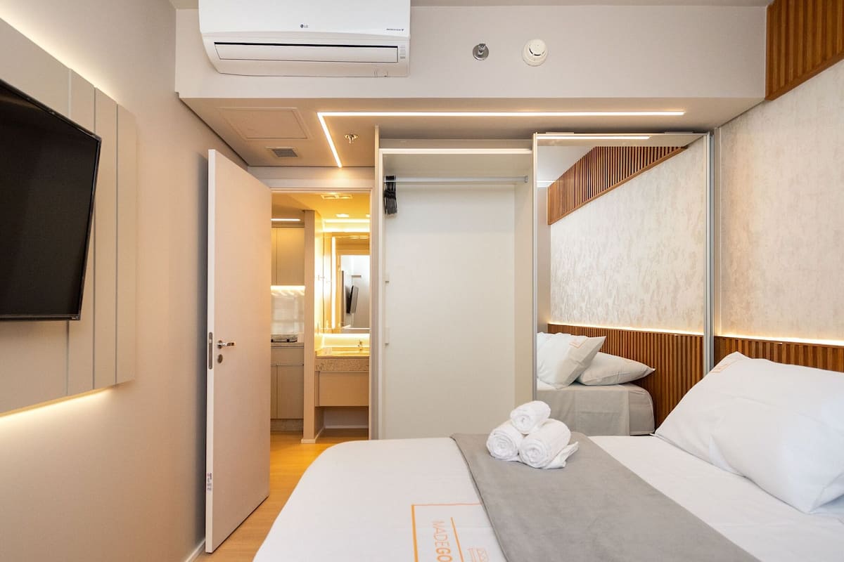 Quarto de hotel com uma cama, televisão, ar condicionado, espelho e banheiro visível por uma porta aberta.