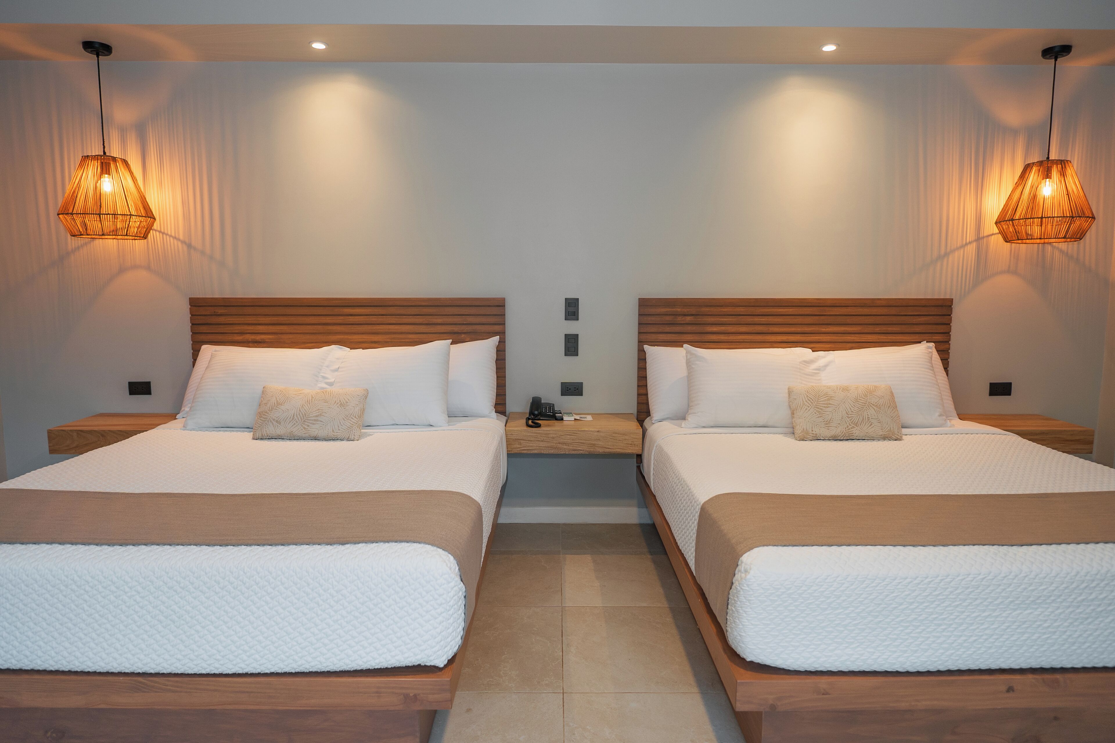 Quarto duplo com duas camas, cada uma com cabeceira de madeira, roupa de cama branca e uma pequena mesa de cabeceira com abajur.