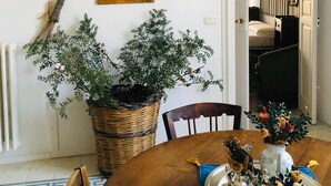 Uma sala de jantar com uma mesa de madeira, cadeiras, um cesto de vime com plantas, um quadro e decorações rústicas nas paredes.
