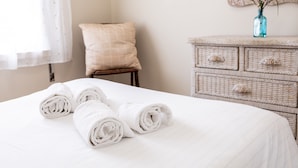 Uma cama bem arrumada com toalhas brancas dobradas, uma cadeira de madeira com almofada e um aparador de vime com gavetas.